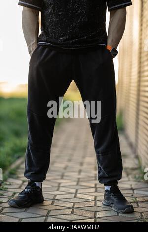 A person in casual clothing stands on a sidewalk holding a phone, with ...