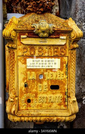A weathered yellow mailbox stands on a metal post behind a barbed wire ...