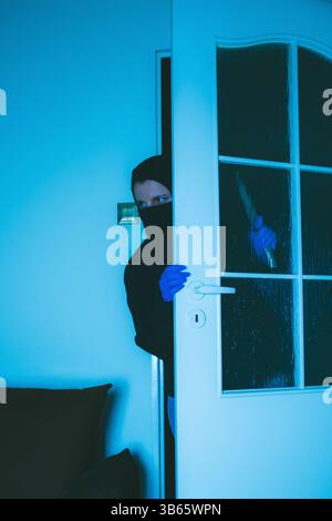 Masked intruder wearing black clothing, holding a knife, peeking ...
