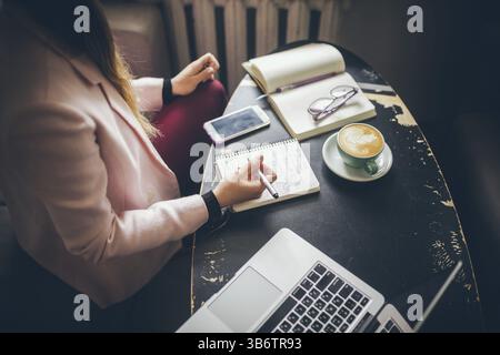 A woman works at a laptop, makes notes in a notebook, planning, routine ...
