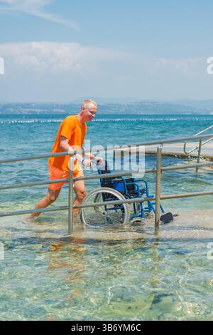 Lifeguard pushing an empty wheelchair from the sea along a beach ramp ...