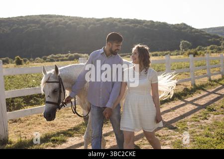 Couple walk at the ranch. The young bearded man leads a white horse ...