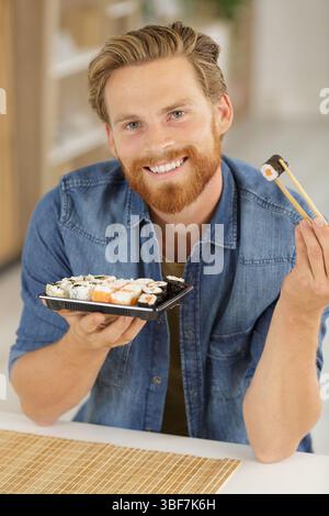Handsome man with beard eating sushi using chopsticks pointing thumb up ...