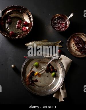 Metal plates with bread, jam, grapes, and a brass fish on dark table ...