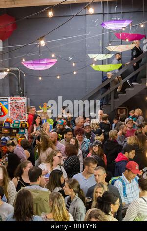 A diverse crowd of people gathered indoors, looking upwards with varied ...