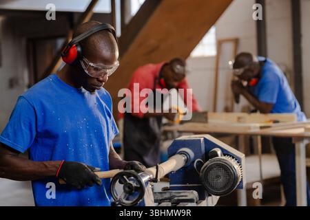 African American male coworkers operating wood lathe and sanding planks ...