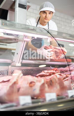Positive young salesman demonstrating piece of meat in butcher shop ...