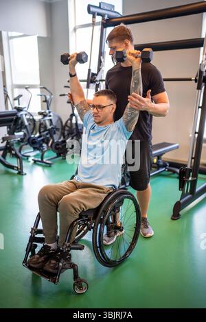 Trainer assists young disabled man in wheelchair lifting dumbbells on ...