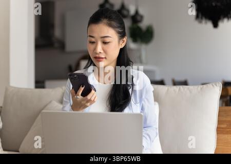 Asian woman using smartphone and laptop computer at home office desk ...