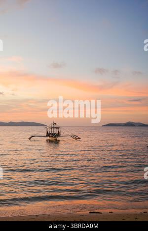 A Filipino boat floats quietly on the calm ocean as the sun sets behind ...