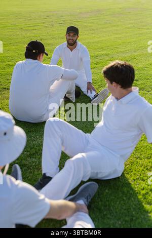 Diverse cricket teammates sitting on outfield chatting in white kit and ...