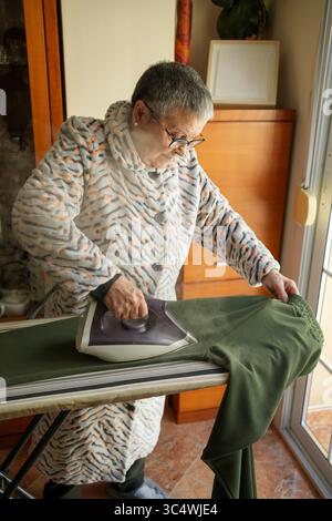 Elderly woman using a steam iron on an ironing board, performing ...