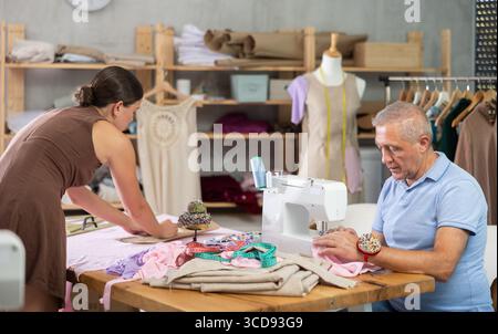 Girl employee sewing, stitch clothes, senior man pins paper pattern to ...