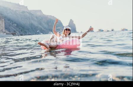 A woman floats on a pink donut inflatable ring in the calm blue sea ...