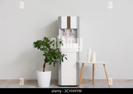 Modern water cooler, houseplant and table with paper cups near white ...