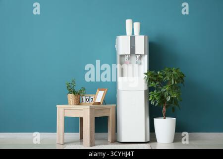 Modern water cooler, houseplant and table with paper cups near white ...