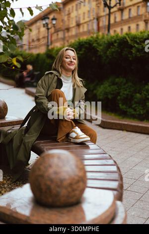 happy woman in trench coat standing near wall and talking on smartphone ...