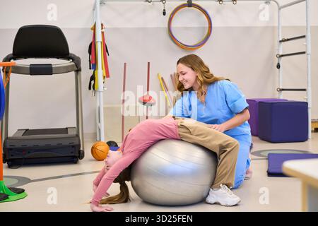 A child exercises on a large stability ball while a friendly therapist ...