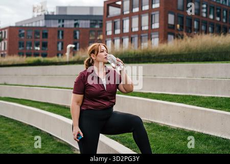 Sporty plus size woman with water bottle sitting on fitness mat at home ...