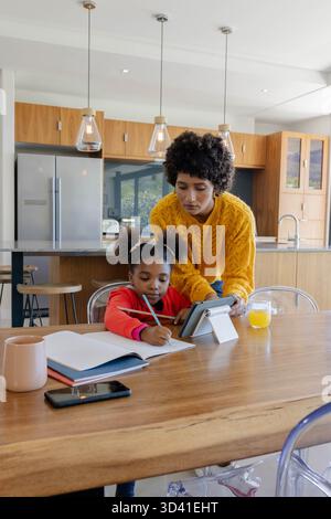 Diverse mother and daughter working at kitchen table, using laptop with ...