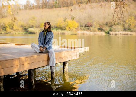 Autumn lake woman. She sits by a pond on a wooden pier in autumn and ...