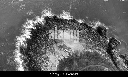 Aerial monotone view of rugged coastal rocks with waves breaking below ...