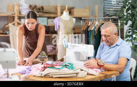 Girl employee sewing, stitch clothes, senior man pins paper pattern to ...