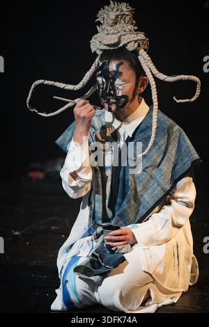 A performance artist sits under dramatic stage light, wearing an ethnic ...