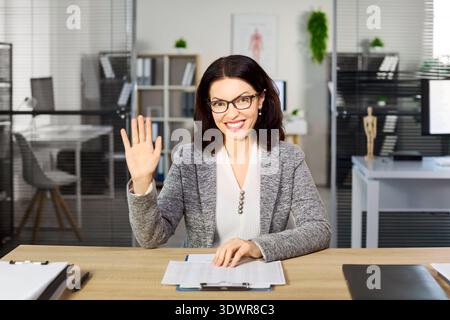 Smiling businesswoman waving during online video call in modern office ...