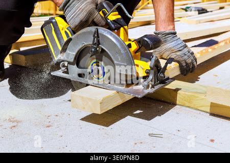 Frame worker operates power hand saw on wooden beam in wooded area ...