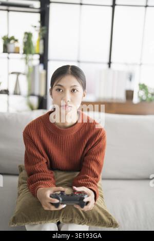 young Asian woman playing video game with controller sitting on sofa at ...