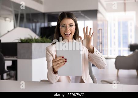 Smiling businesswoman waving during online video call in modern office ...
