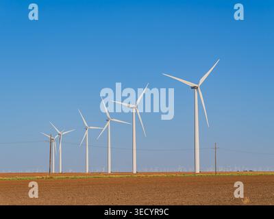 White wind turbines stand in a harvested field under a blue sky with ...