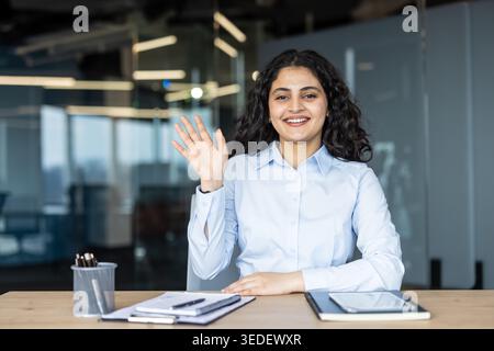 Smiling businesswoman waving during online video call in modern office ...
