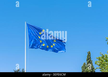 A large European Union flag waves on a tall white pole against a clear ...