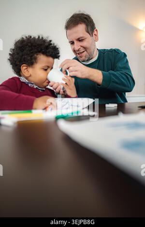 Father helping his son drink from a mug while they are drawing at home ...