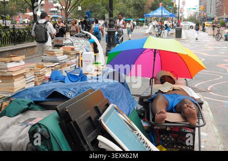 A person sleeps under an umbrella next to piles of books stacked on a ...