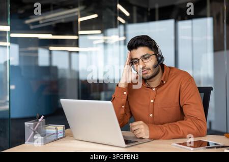 Man with headset and glasses sits at laptop in bright office, speaking ...