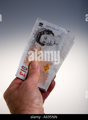A hand holding a fan of twenty-pound banknotes, with stacks of more ...