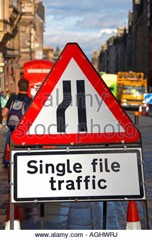 A triangular roadworks sign and traffic cone at the side of a road at ...