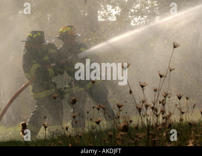 Two firefighters on a hoseline spraying water into a fire surrounded by ...