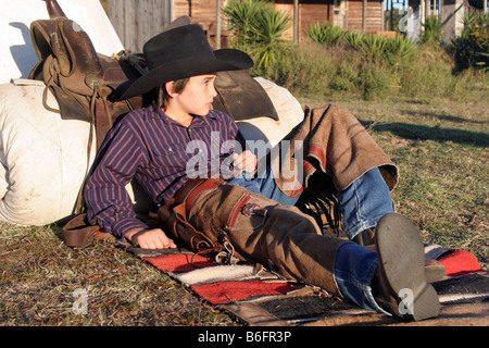 A young cowboy resting against his bed roll and saddle out on the range ...