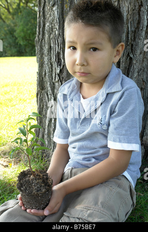 Six year old boy holds small tree, seated beside large tree, Winnipeg ...
