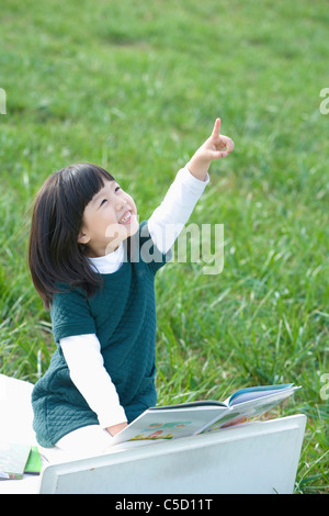 Girl sitting reading a book, pointing at the page with toy lamb beside ...