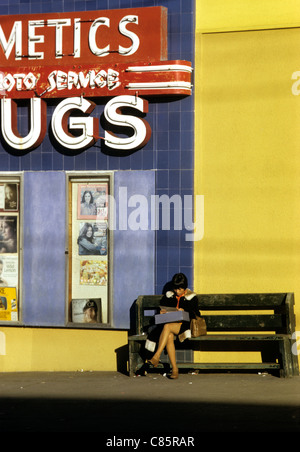 A woman sits at a bus stop as a city worker sweeps the street in ...