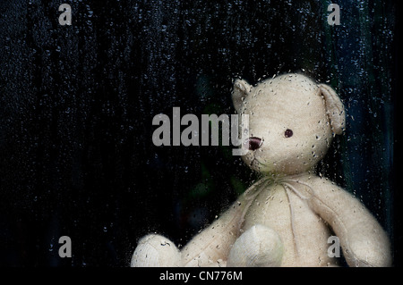 Sad Teddy bear looking through a window covered in rain drops. Still ...