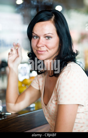 young woman smoking cigarette while sitting at wooden table with camera ...