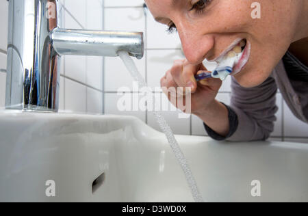 Woman is brushing her teeth, while water is still running from the tap ...
