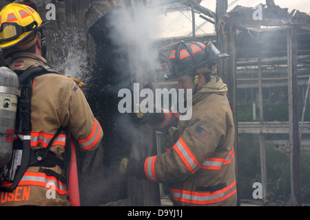 Two firefighters on a hoseline spraying water into a fire surrounded by ...