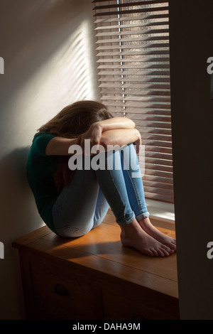 A teenage girl, head between her knees, sitting by a window with light ...